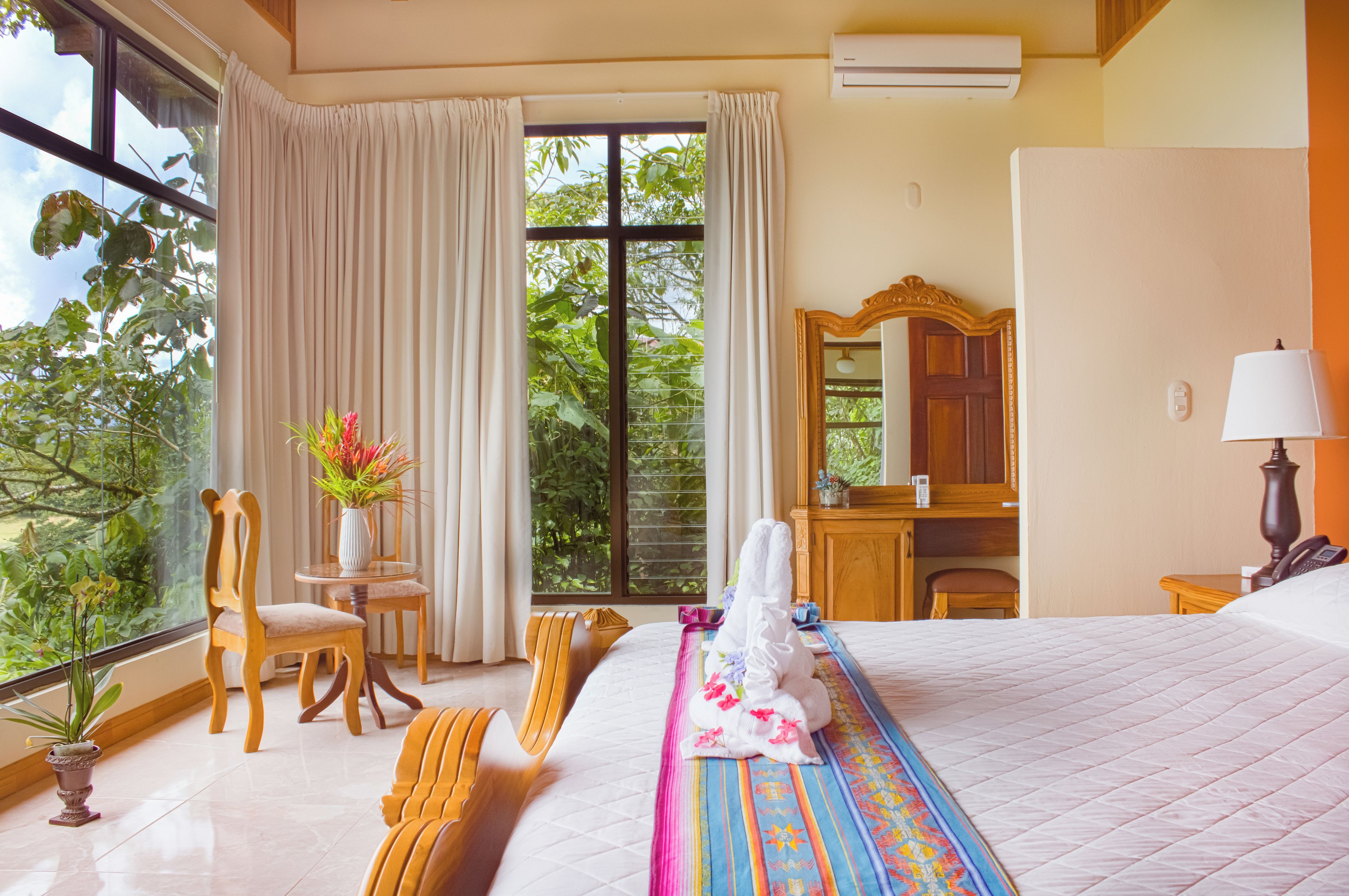 Quarto de hotel com cama, escrivaninha, cadeira, janela com cortinas, espelho e mesa de cabeceira com abajur.