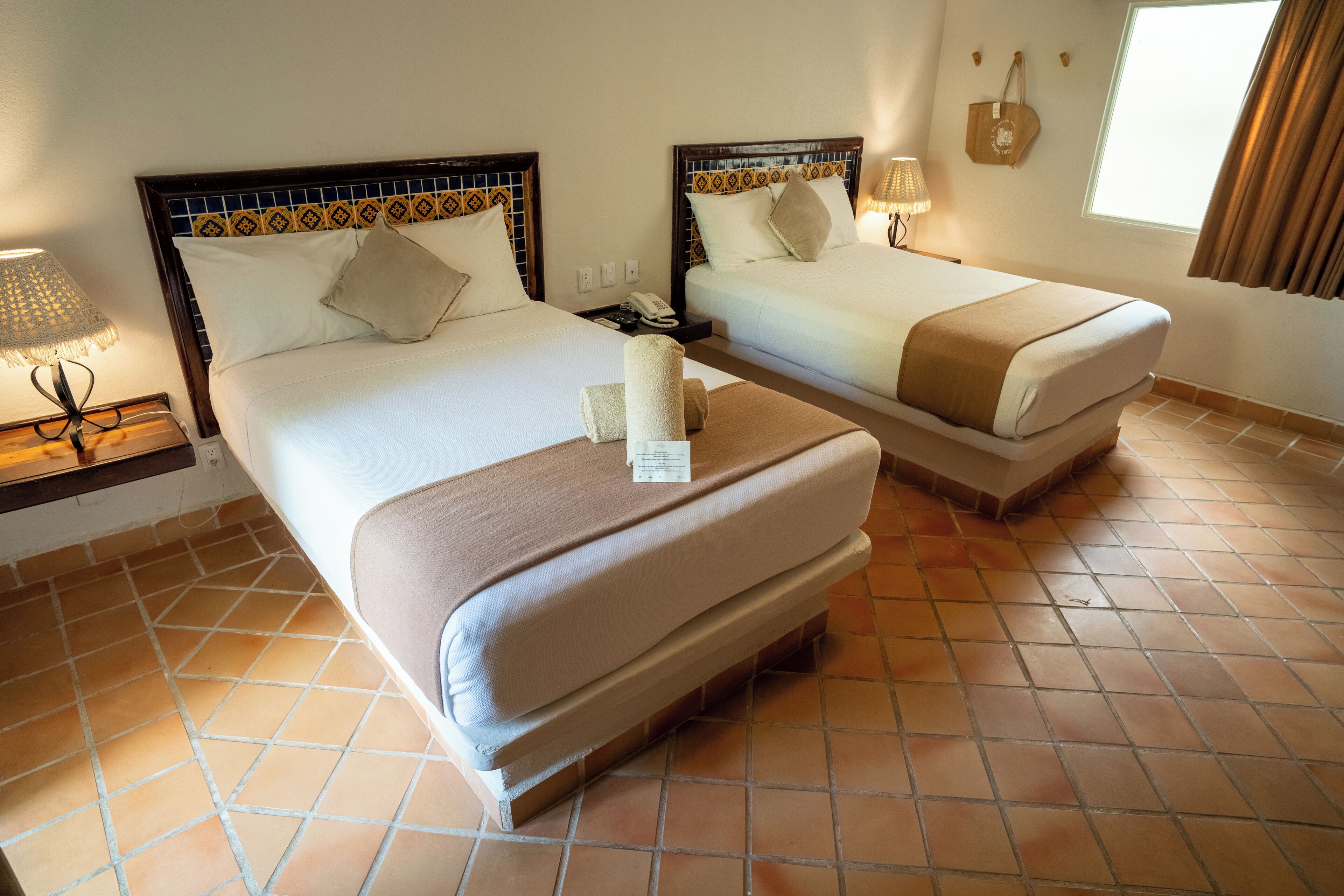 Quarto de hotel com duas camas, chão de azulejos, mesa de cabeceira com abajur e janela com cortinas.