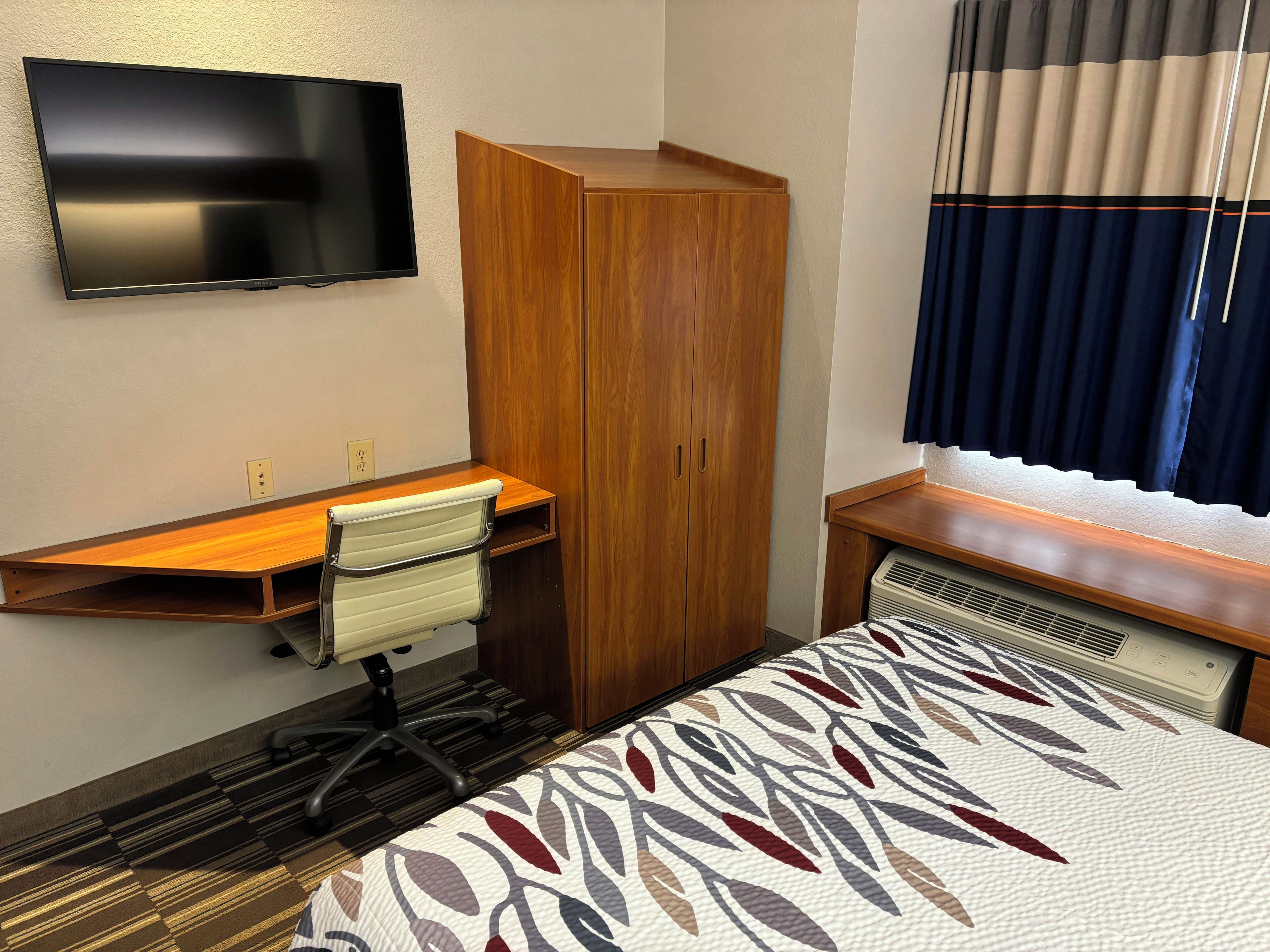 Quarto de hotel com cama, escrivaninha com cadeira, guarda-roupa, televisão na parede e janela com cortinas.