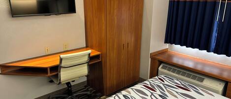 Quarto de hotel com cama, escrivaninha com cadeira, guarda-roupa, televisão na parede e janela com cortinas.