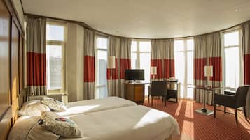 Quarto de hotel com duas camas, uma escrivaninha, uma cadeira e janelas amplas com cortinas.