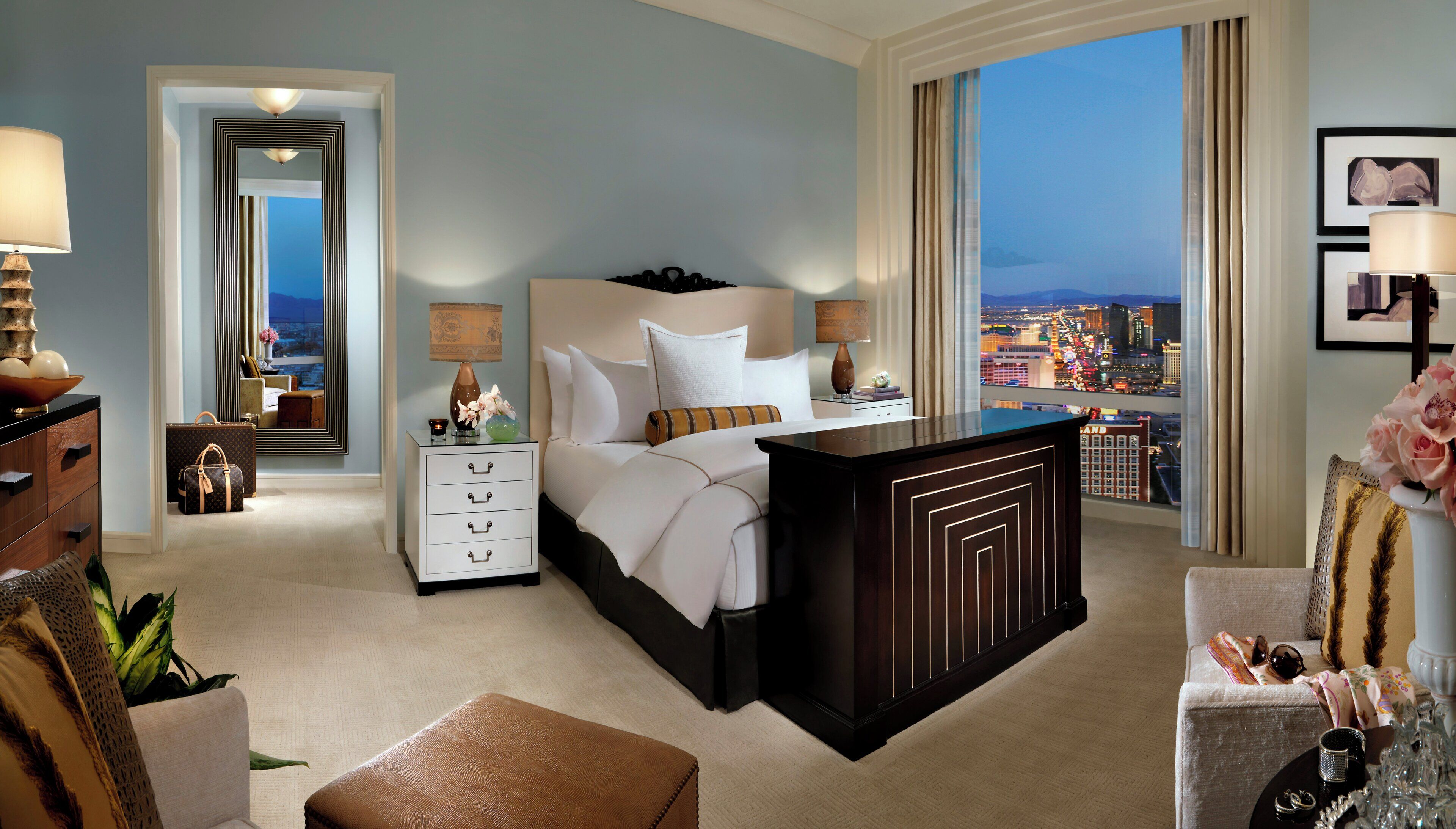Quarto de hotel com uma cama grande, mesa de cabeceira, cômoda, área de estar com uma poltrona e repouso para os pés, e vista para a paisagem urbana.