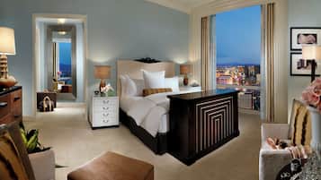 Quarto de hotel com uma cama grande, mesa de cabeceira, cômoda, área de estar com uma poltrona e repouso para os pés, e vista para a paisagem urbana.