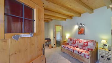 Interior em madeira com porta de madeira, sofá com capa estampada em vermelho e branco, pequeno armário de madeira com gavetas e luminária fixada na parede.