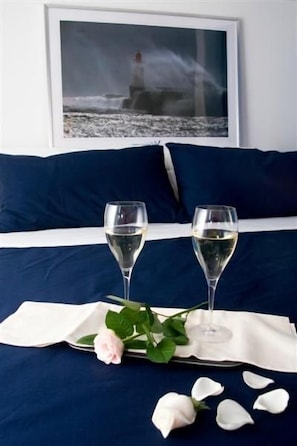 Uma cama com dois copos de vinho branco, uma rosa e pétalas de flores brancas sobre um edredom azul marinho.