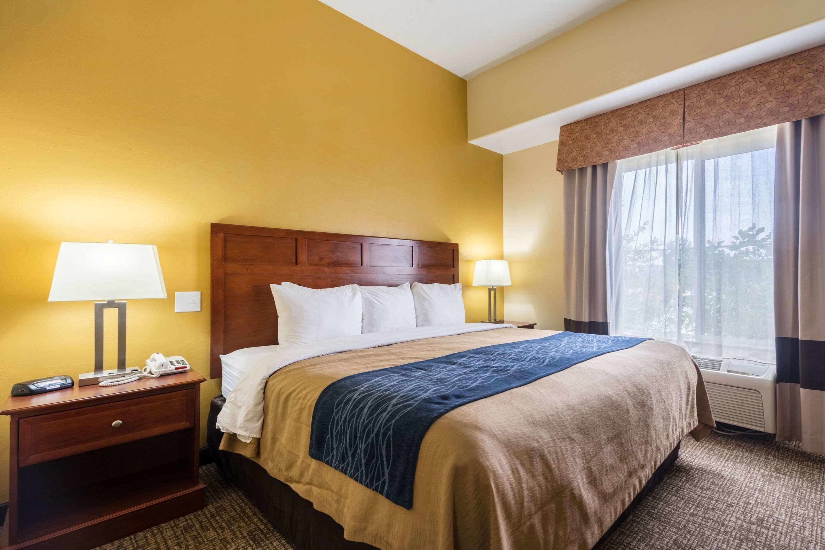 Quarto de hotel com uma cama grande, duas luminárias laterais, uma mesa de cabeceira de madeira com telefone e uma janela com cortinas.