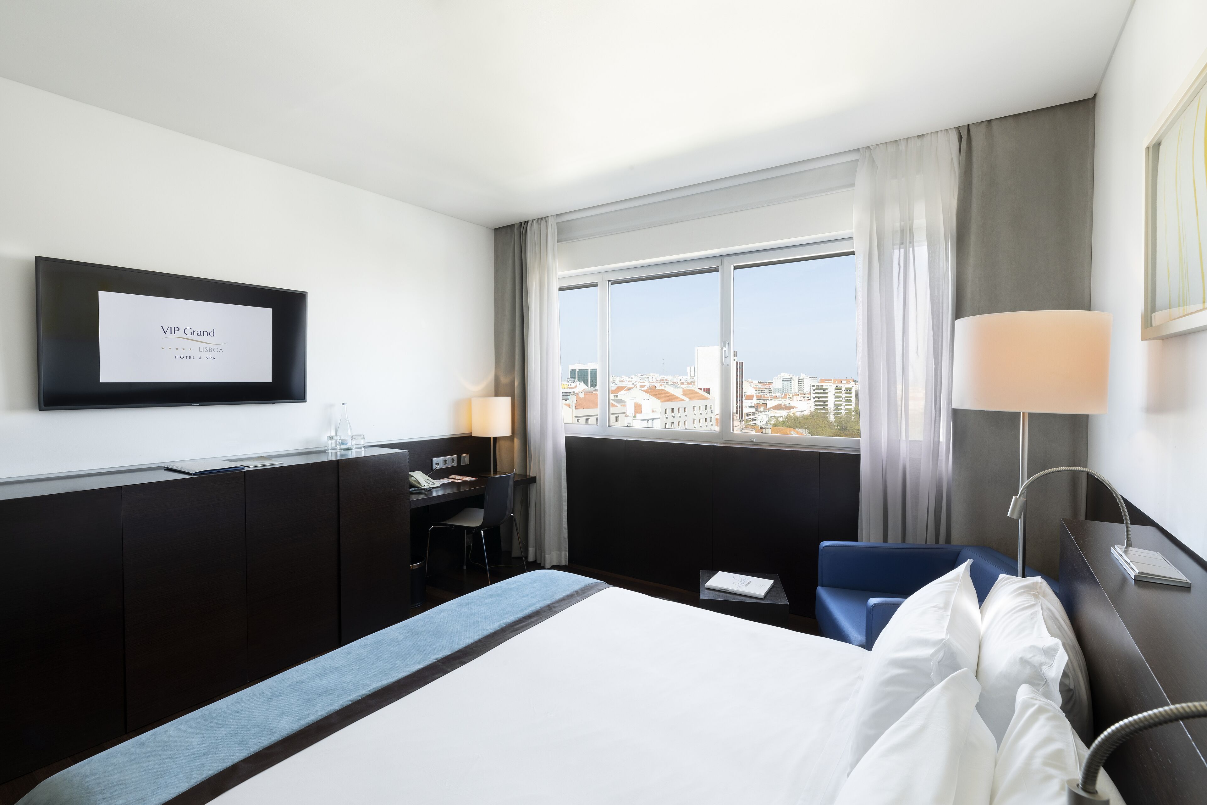 Quarto de hotel com cama, escrivaninha, cadeira, abajur e vista da cidade.