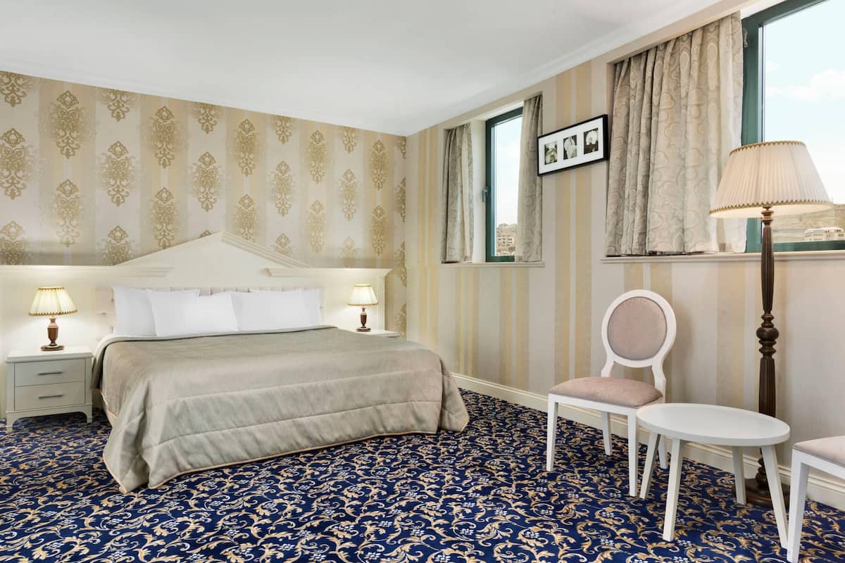 Quarto de hotel com uma cama grande, duas mesas de cabeceira com luminárias, uma cadeira, uma mesinha e uma janela com cortinas.