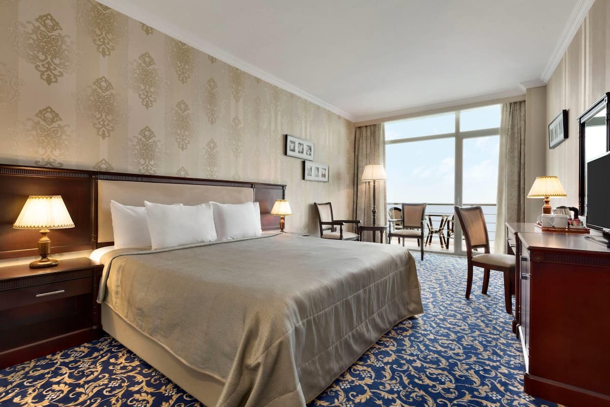 Quarto de hotel com uma cama grande, duas mesas de cabeceira com luminárias, uma escrivaninha com cadeira e vista para o exterior através de uma grande janela.