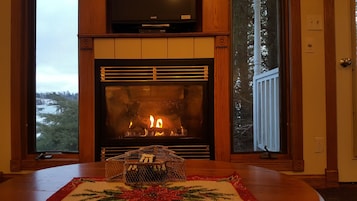 Lareira com televisão acima, mesa com objeto decorativo e vista para árvores lá fora.