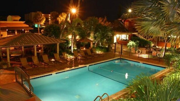 Uma área de piscina à noite, com espreguiçadeiras e guarda-sóis, cercada por palmeiras e edifícios.