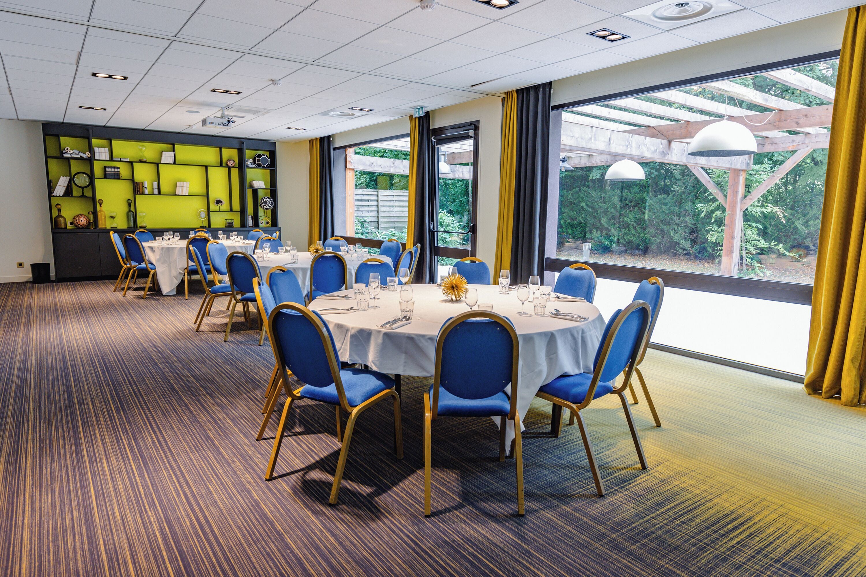 Sala de conferências com mesas redondas, cadeiras azuis e vista para o verde através de grandes janelas.