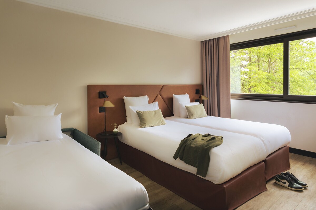 Quarto de hotel com duas camas, uma mesa de cabeceira, uma luminária e uma janela com vista para árvores.