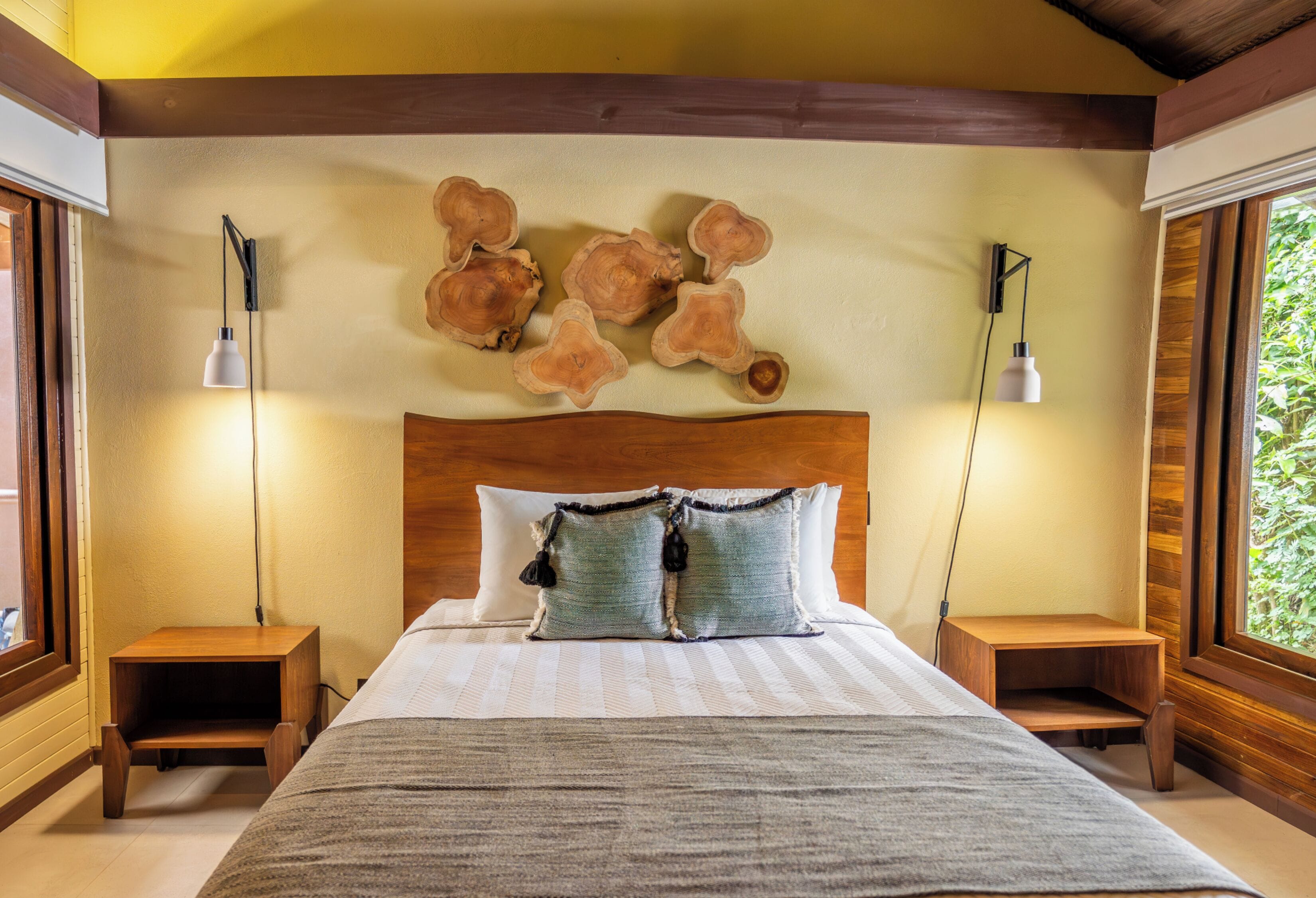 Quarto com cabeceira de madeira, duas mesas de cabeceira, cama com roupa de cama listrada e luminárias fixadas na parede.
