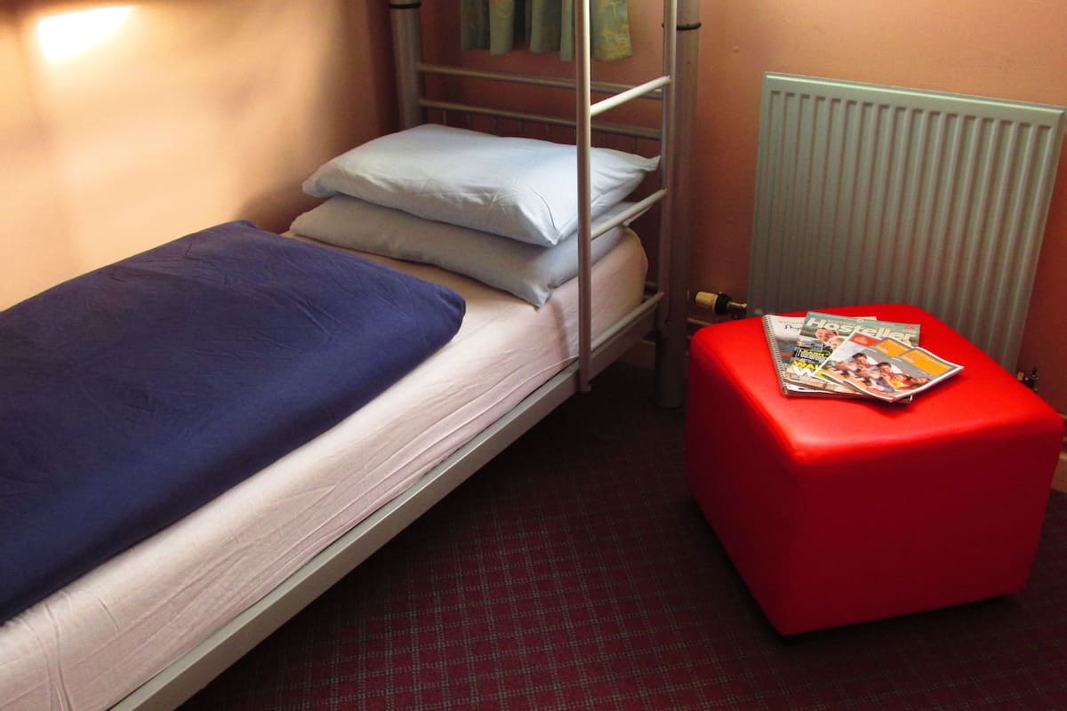 Uma cama de solteiro com um cobertor azul, um banquinho vermelho e uma revista sobre o banquinho.
