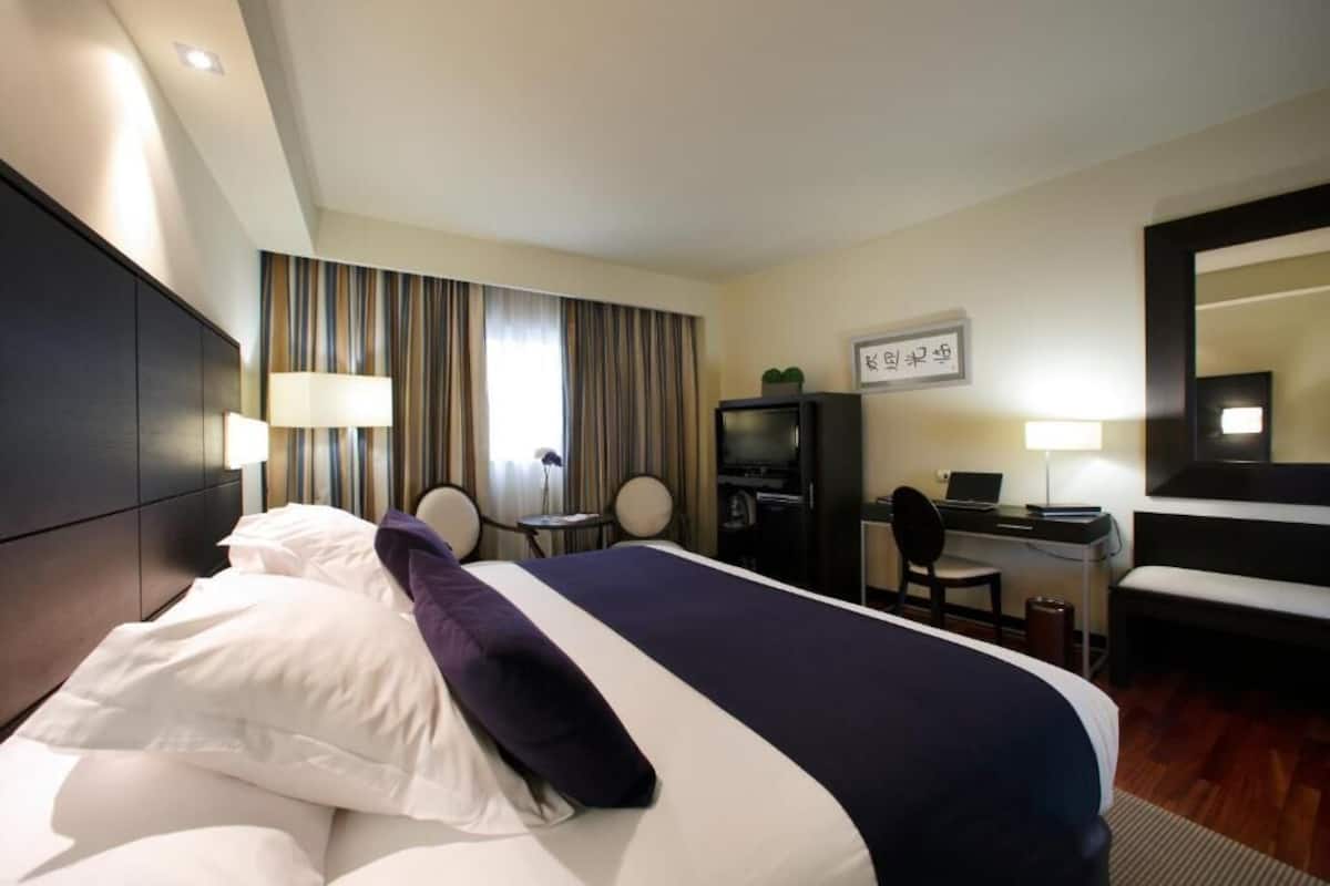 Quarto de hotel com uma cama grande, uma escrivaninha, uma cadeira, uma televisão e uma luminária.
