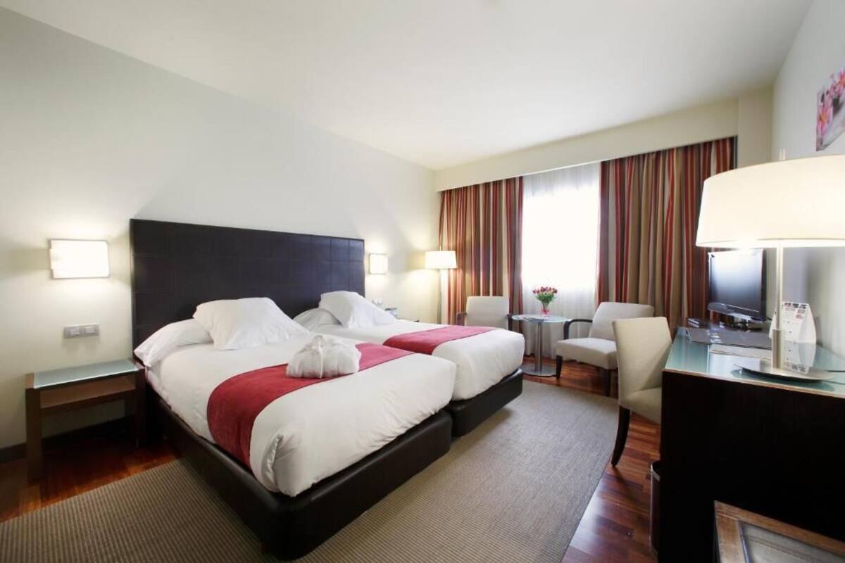 Quarto de hotel com duas camas, uma escrivaninha, uma cadeira, uma televisão e um abajur.