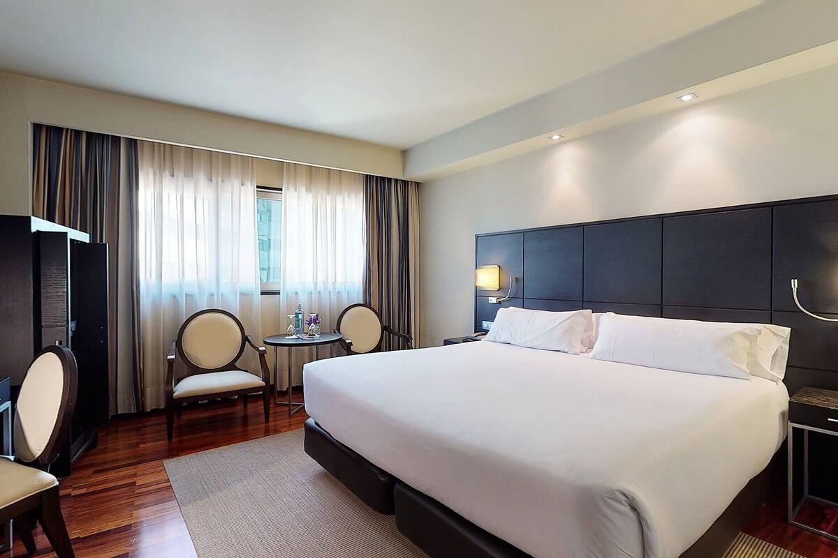 Quarto de hotel com cama, duas cadeiras, escrivaninha, mesinha e criado-mudo com abajur.