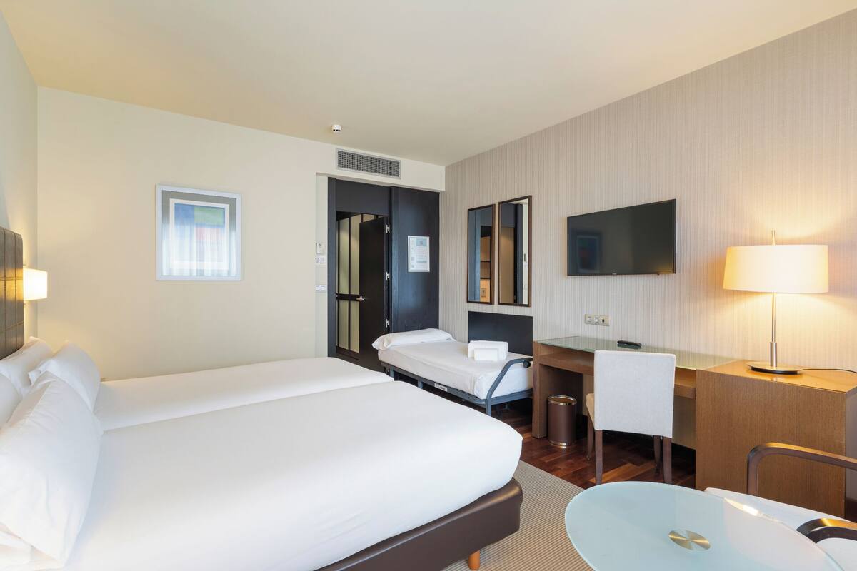 Quarto de hotel com uma cama grande, uma escrivaninha, uma cadeira, uma televisão e um abajur.