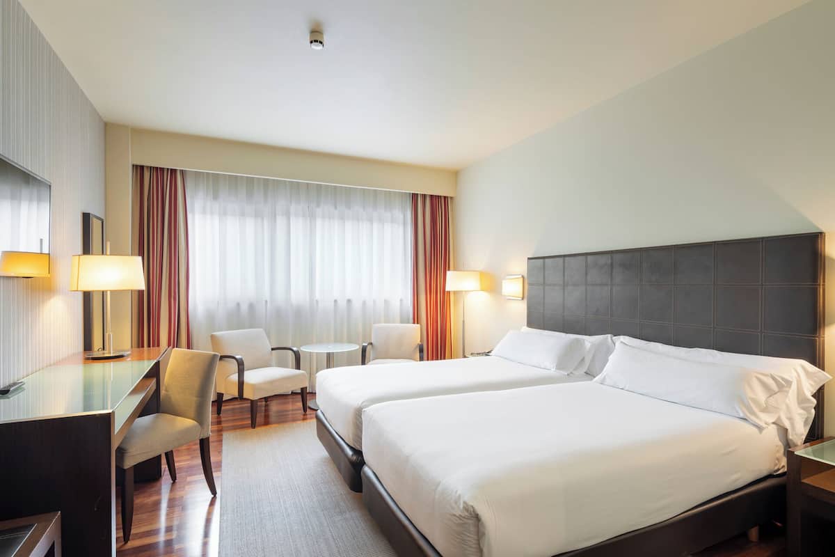 Quarto de hotel com duas camas, uma escrivaninha, uma cadeira, uma mesinha e uma janela grande com cortinas.