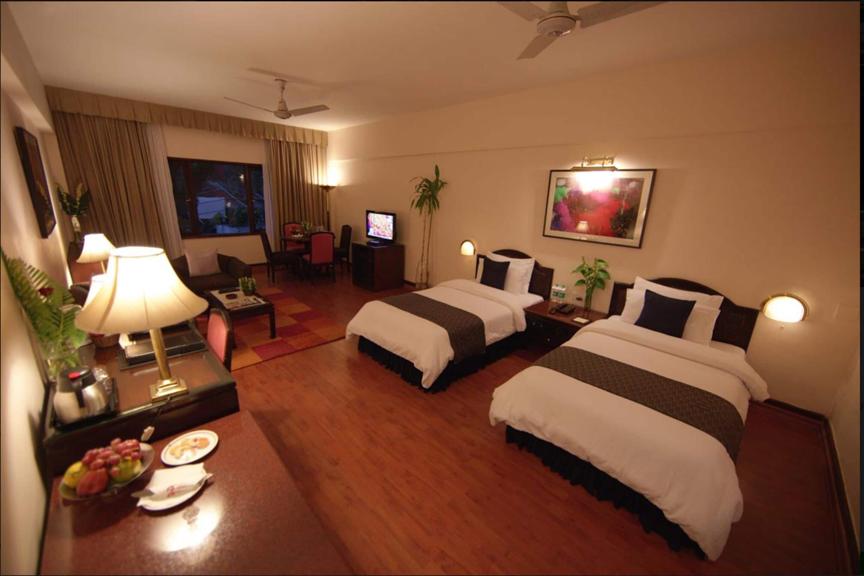 Quarto de hotel com duas camas, uma mesa de escritório, uma televisão e um ventilador de teto.
