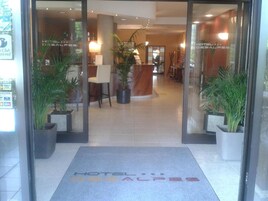 Entrada do Hotel Des Alpes, com balcão de recepção, cadeiras e plantas em vasos.