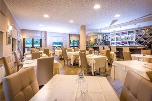Um restaurante com mesas preparadas para refeições, um espaço de bar com prateleiras de garrafas e vista para o exterior através de grandes janelas.
