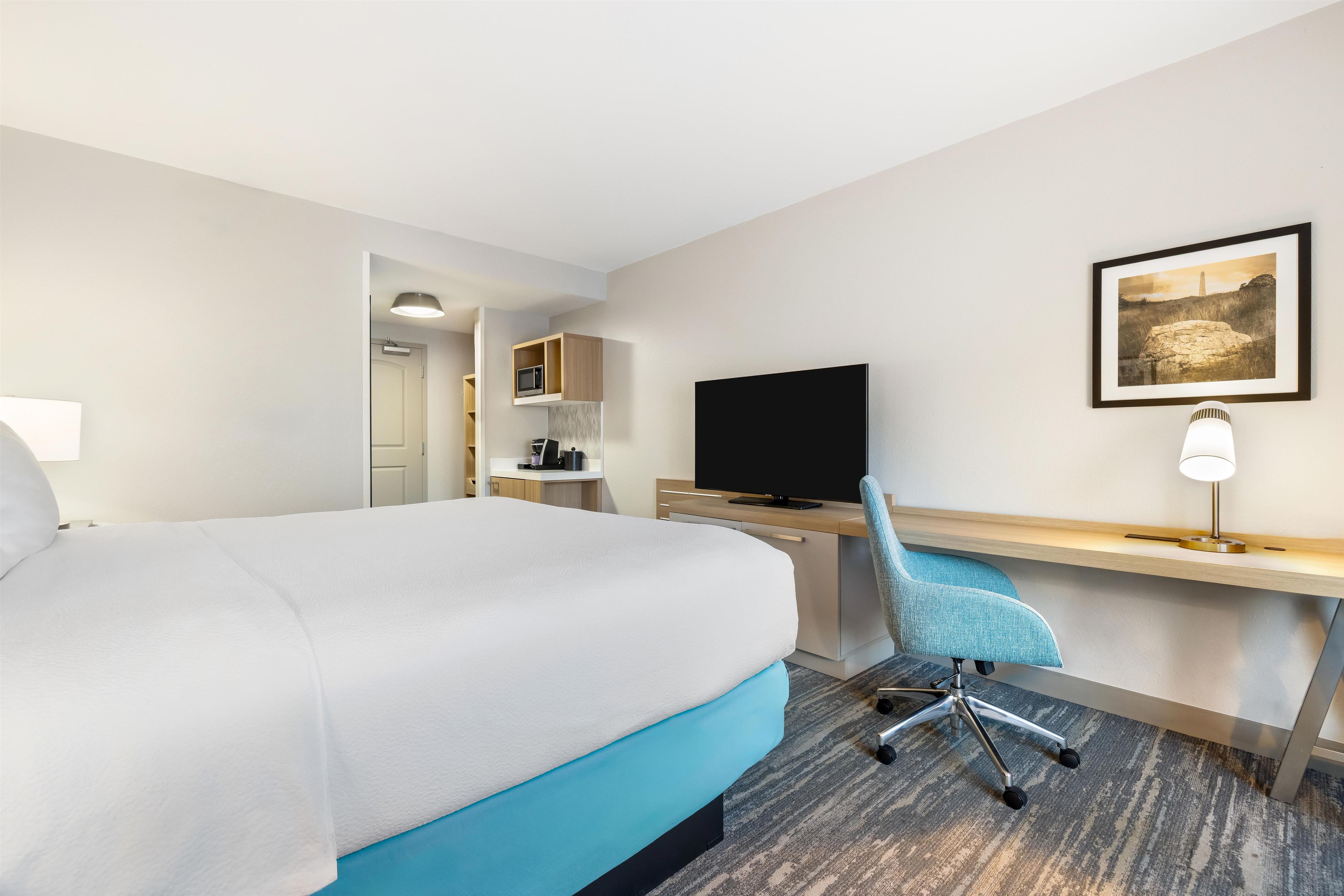 Quarto de hotel com cama, escrivaninha com computador, cadeira, televisão e quadro na parede.