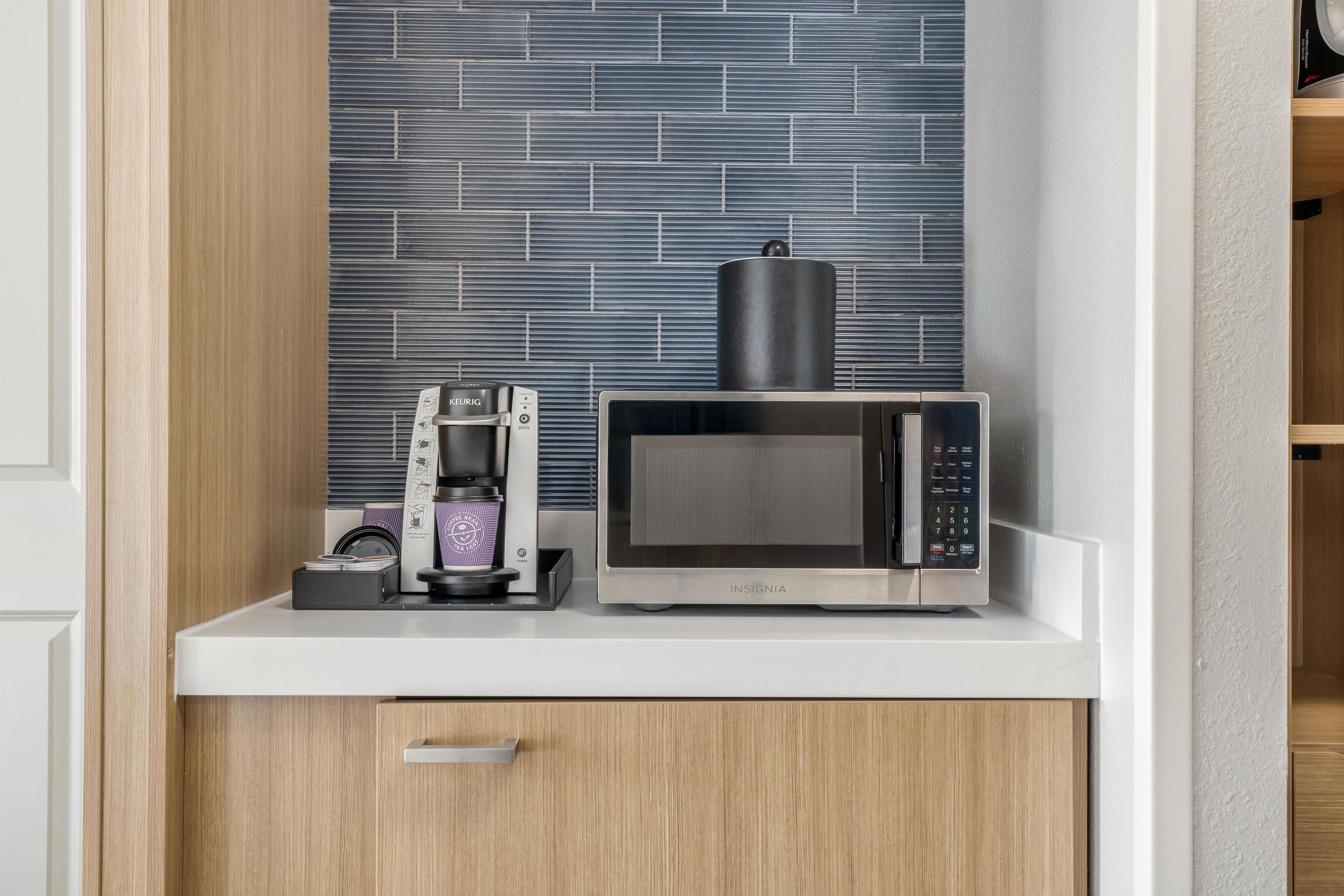 Cozinha compacta com cafeteira, micro-ondas e suporte para cápsulas de café em bancada branca.