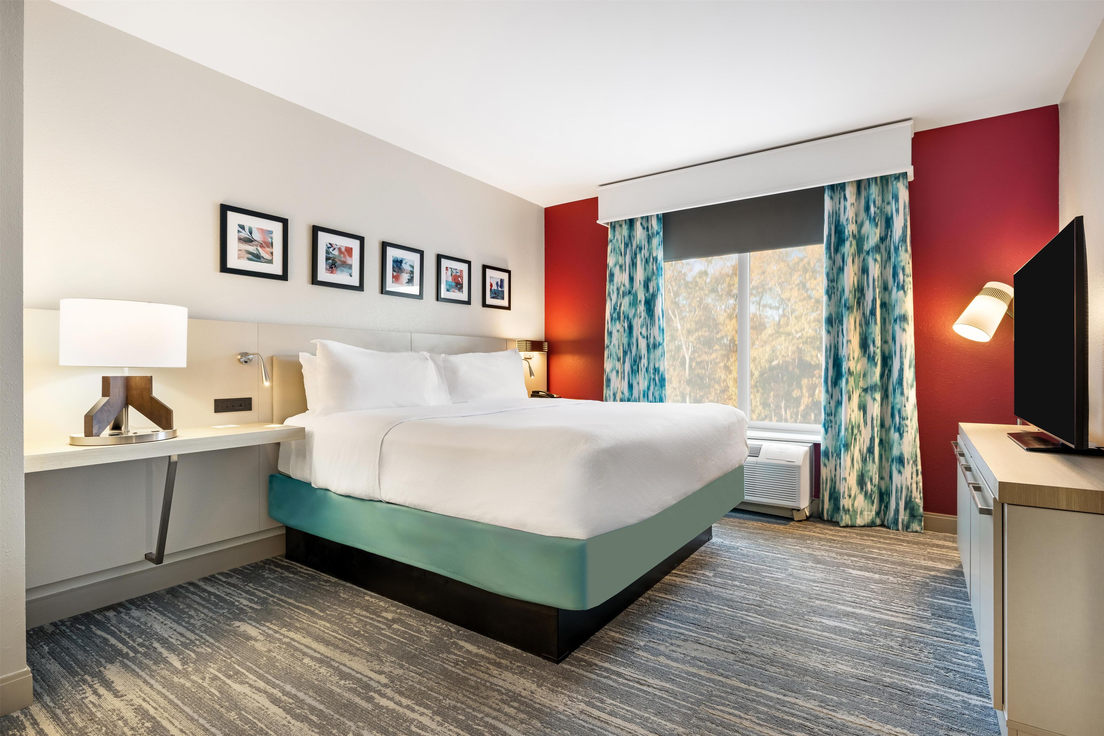 Quarto de hotel com cama, mesas de cabeceira, televisão, janela com cortinas e quadros na parede.