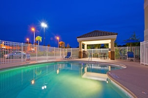 Piscina com área de estar coberta, cercada por um muro branco e iluminada por postes de luz.