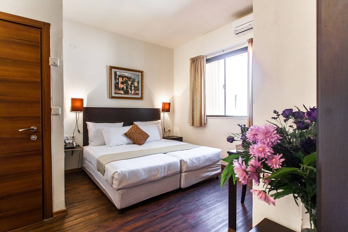 Quarto de hotel com cama, abajures, um quadro na parede, janela com cortinas e um arranjo floral.