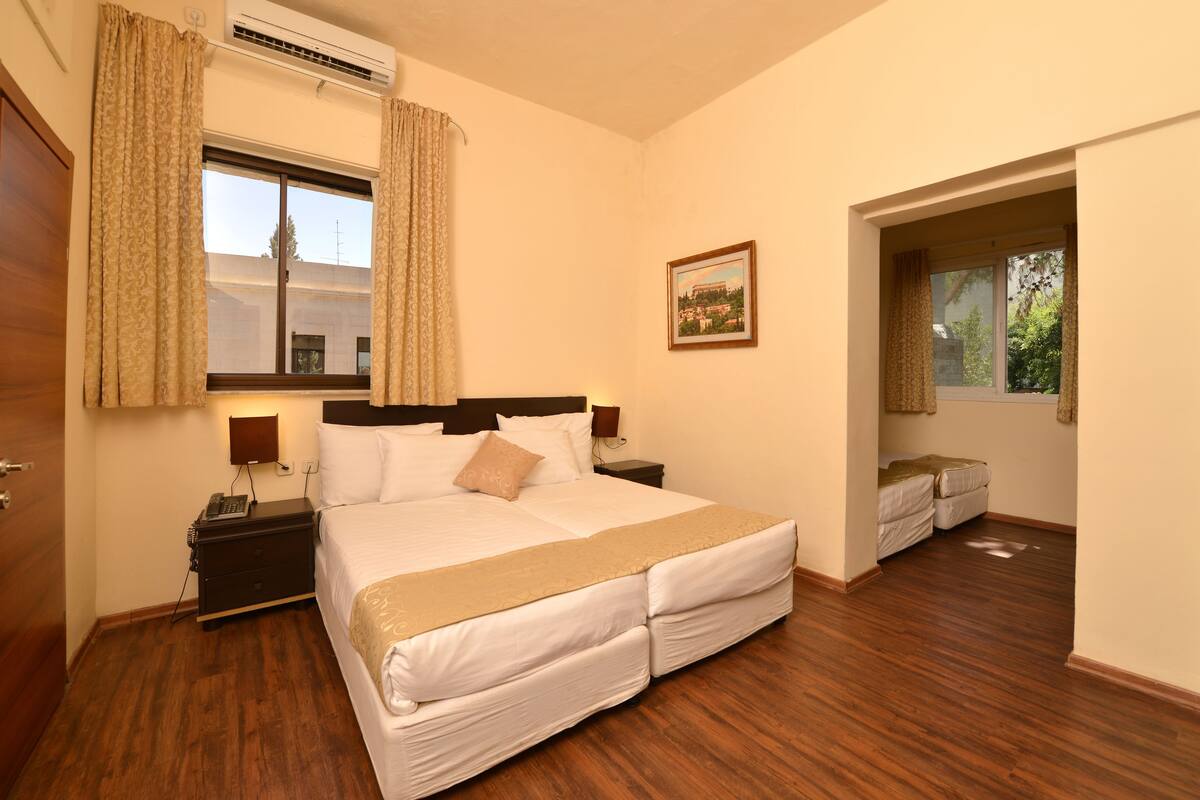 Quarto de hotel com uma cama grande, mesa de cabeceira com abajur, aparelho de ar condicionado e janela com cortinas.