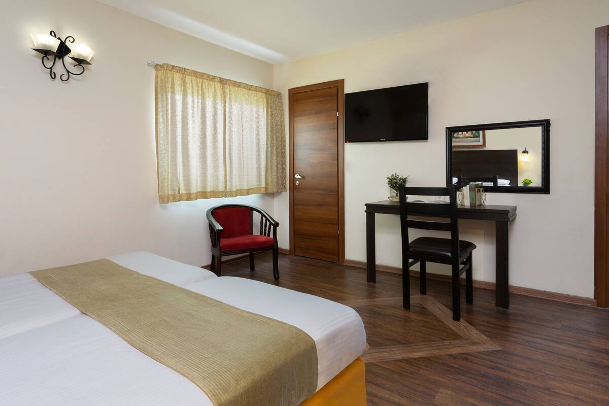 Quarto de hotel com cama, escrivaninha, cadeira, televisão e janela com cortinas.