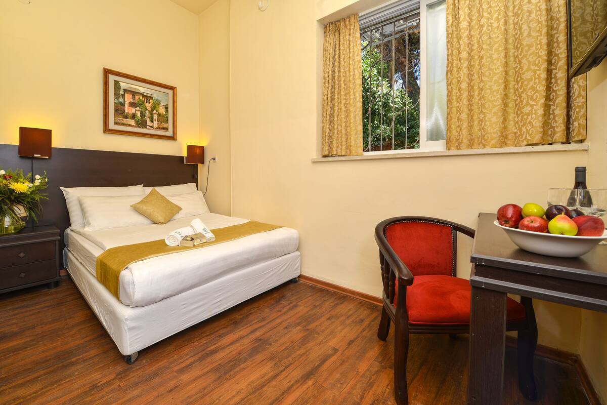 Quarto de hotel com cama, escrivaninha com uma cesta de frutas, cadeira, mesa de cabeceira, luminária e janela com cortinas.