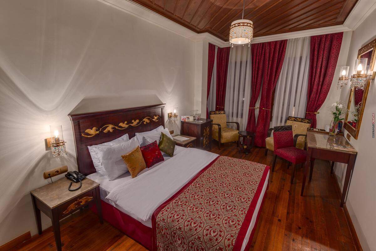 Quarto de hotel com teto de madeira, cama grande com roupa de cama branca e vermelha, escrivaninha com espelho e lustre.