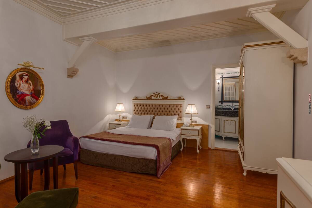 Quarto de hotel com uma cama grande, duas mesas de cabeceira com abajures, uma poltrona roxa, uma pequena mesa redonda com um vaso e um quadro na parede.