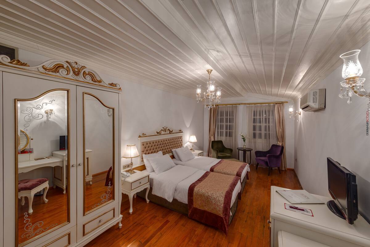 Quarto de hotel com um espelho grande, uma cama com duas roupas de cama brancas, piso de madeira, uma televisão e uma escrivaninha com uma cadeira.
