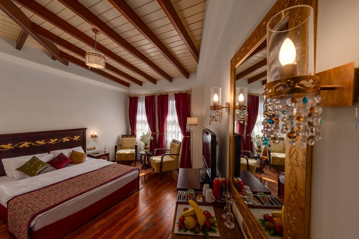 Quarto de hotel com uma cama grande, vigas de madeira no teto, televisão, espelho e uma mesa de jantar com frutas.