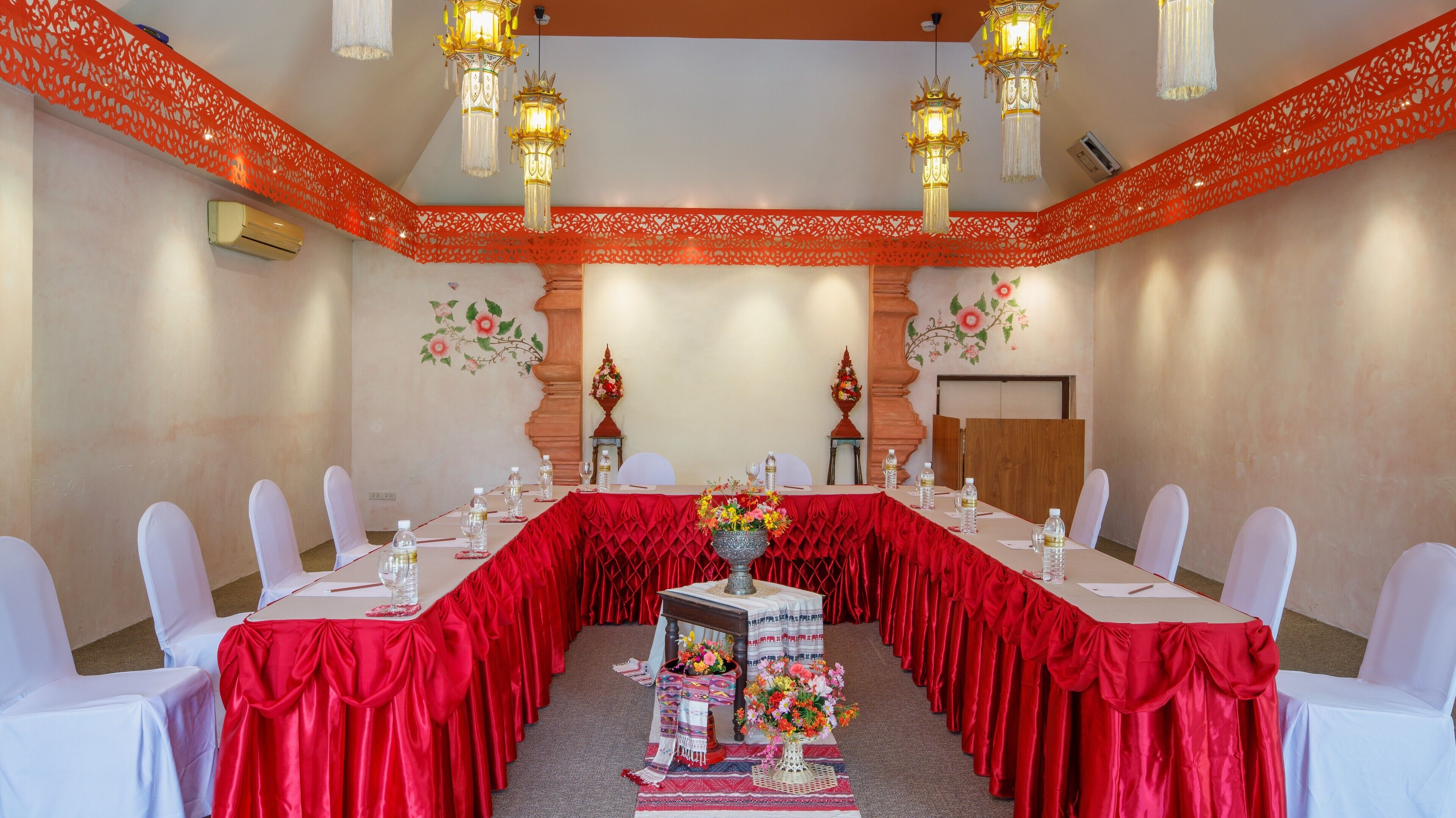 Uma sala de conferências com uma mesa comprida, cadeiras brancas, toalhas de mesa vermelhas e elementos decorativos como lanternas penduradas e arte floral na parede.