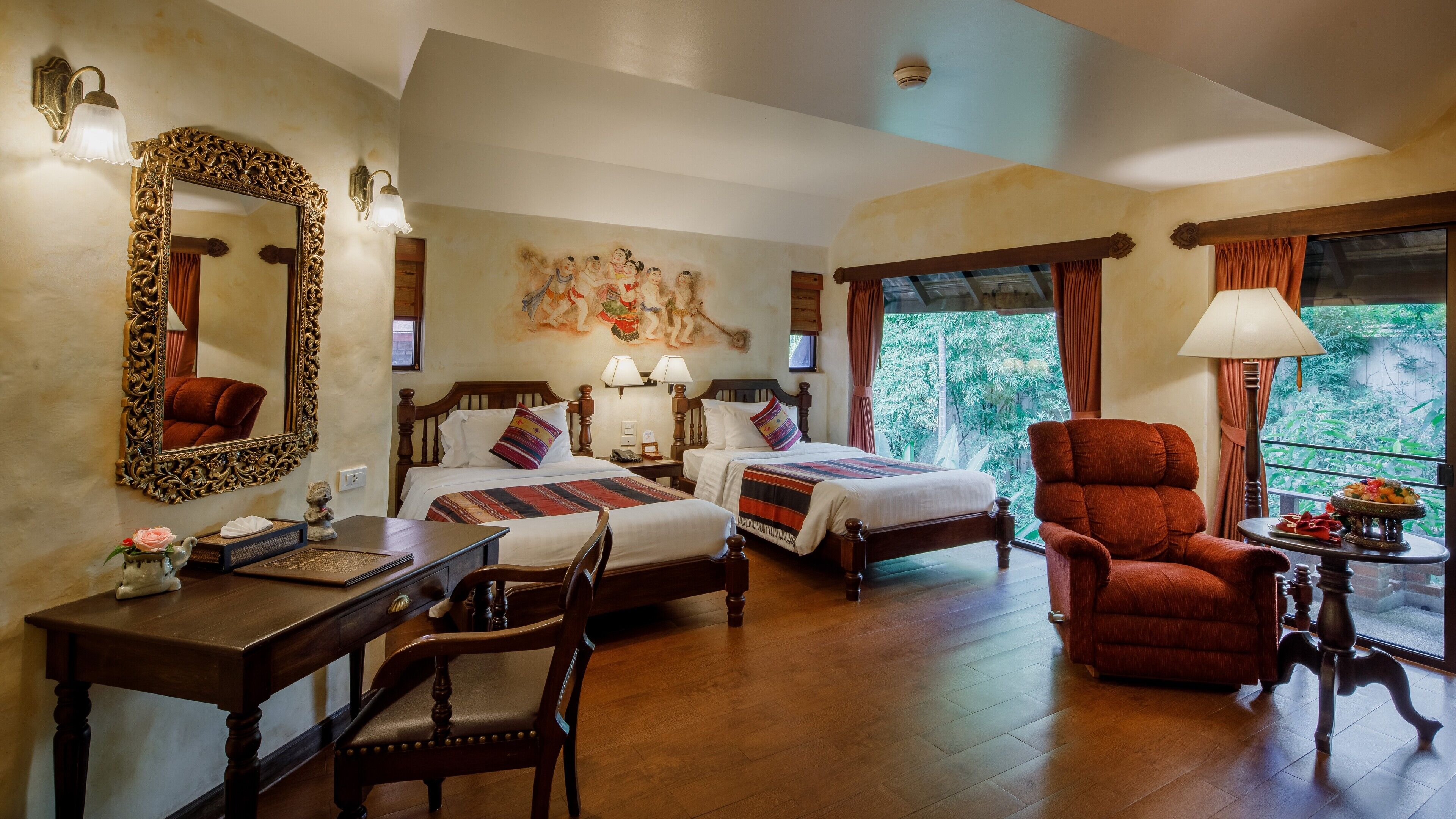 Quarto de hotel com duas camas, uma escrivaninha, uma cadeira, uma poltrona vermelha, uma mesa com uma fruteira e uma janela grande com vista para o verde.