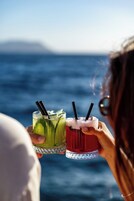 Duas pessoas segurando copos com bebidas e canudos, um verde e um vermelho, com um corpo de água e uma massa de terra ao fundo.