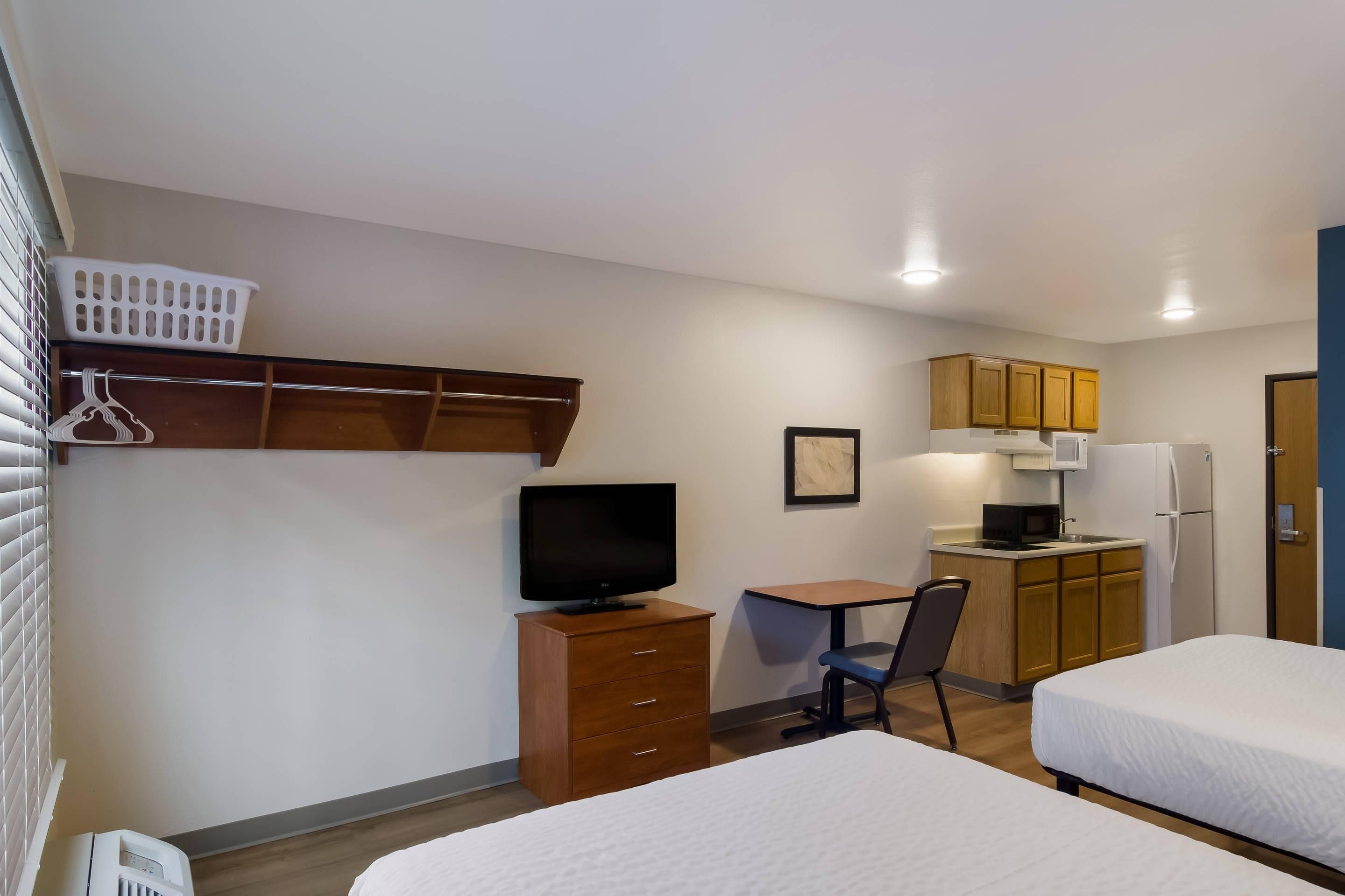 Quarto de hotel com cama, escrivaninha, cadeira, televisão, bancada com micro-ondas e guarda-roupa.