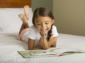 Criança deitada numa cama com um livro, em um quarto com colcha branca e cabeceira de madeira.