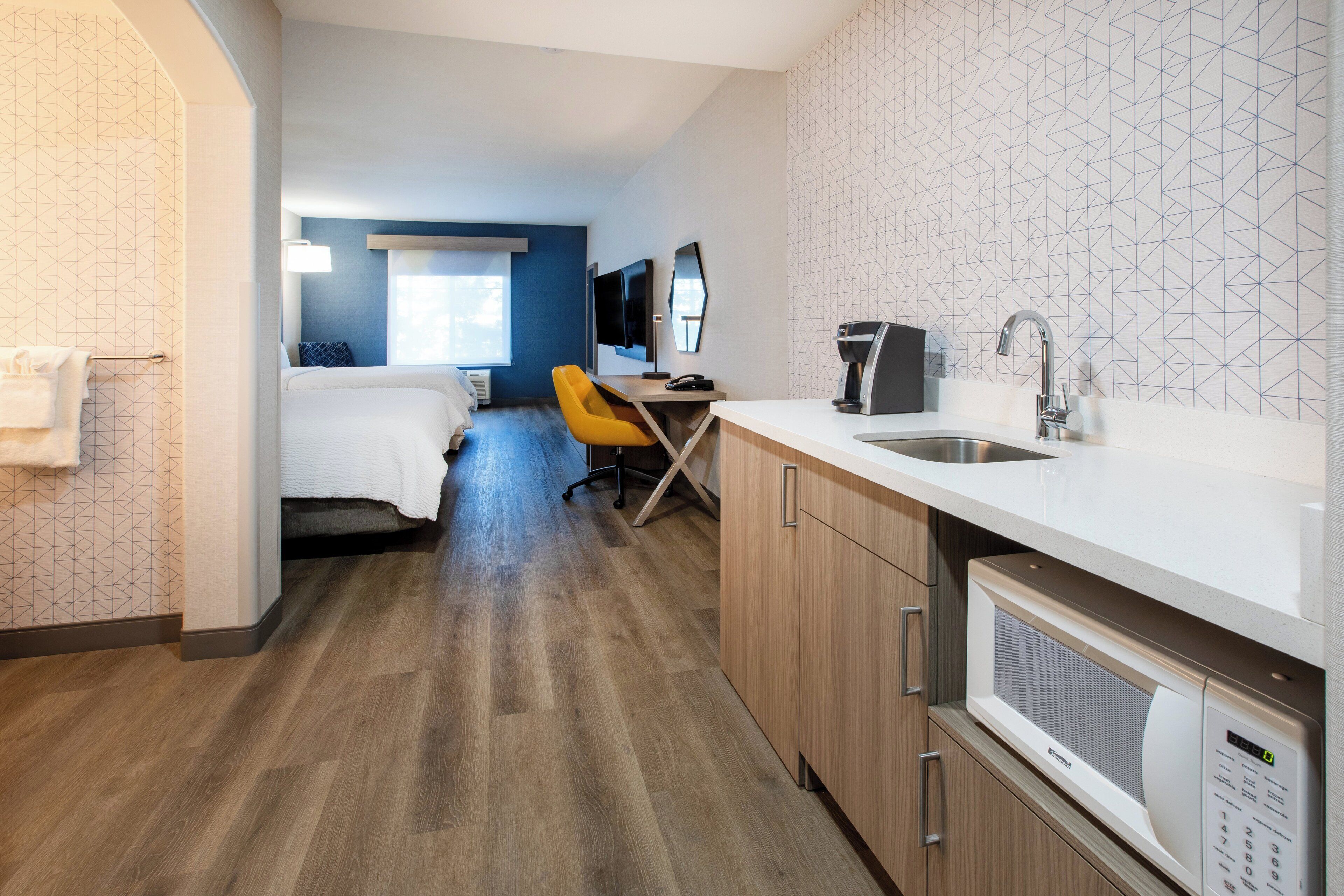 Quarto de hotel moderno com piso de madeira, uma cama, uma escrivaninha com cadeira, televisão e micro-ondas.