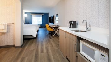 Quarto de hotel moderno com piso de madeira, uma cama, uma escrivaninha com cadeira, televisão e micro-ondas.