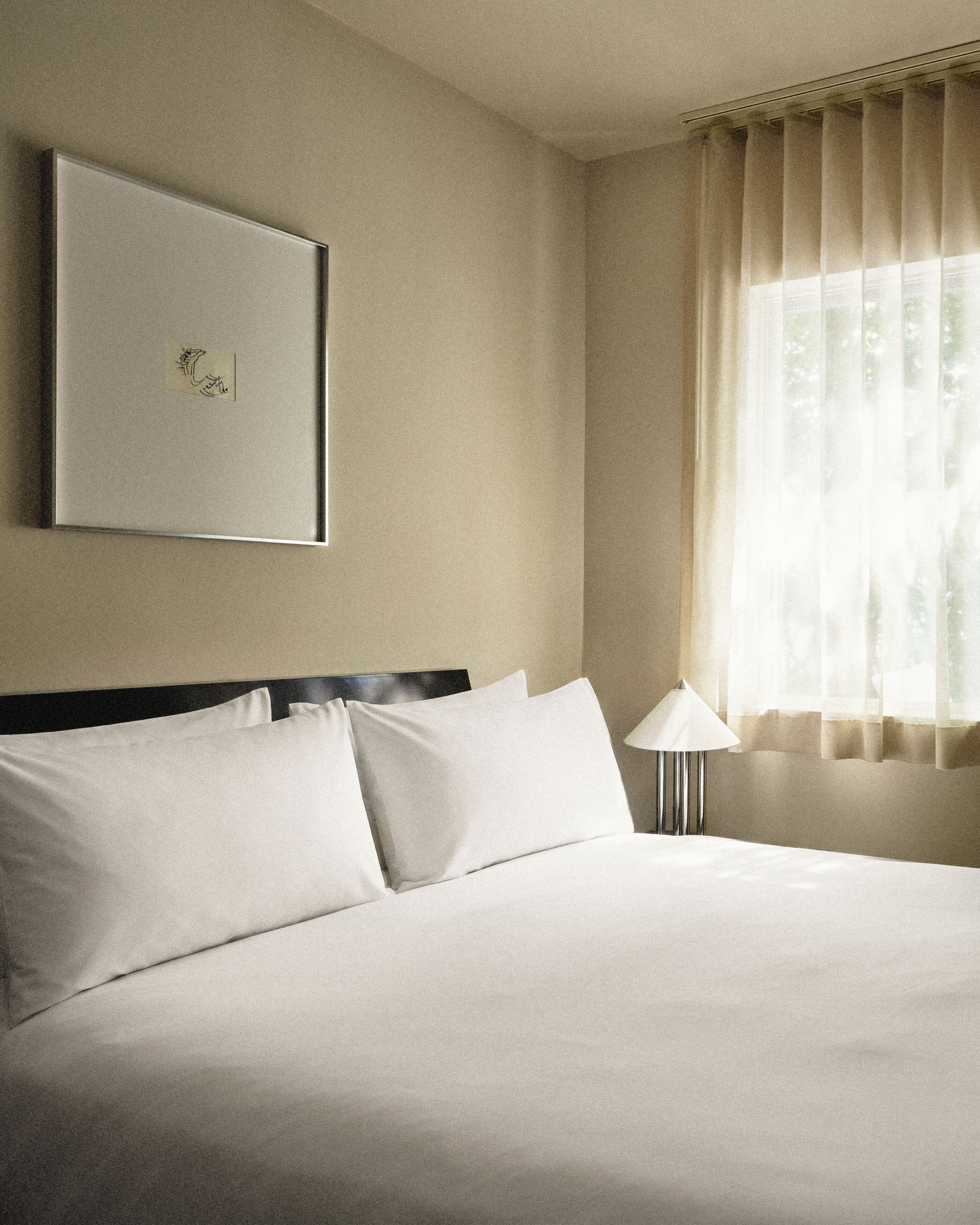 Quarto de hotel com cama, luminária, quadro na parede e janela com cortinas.