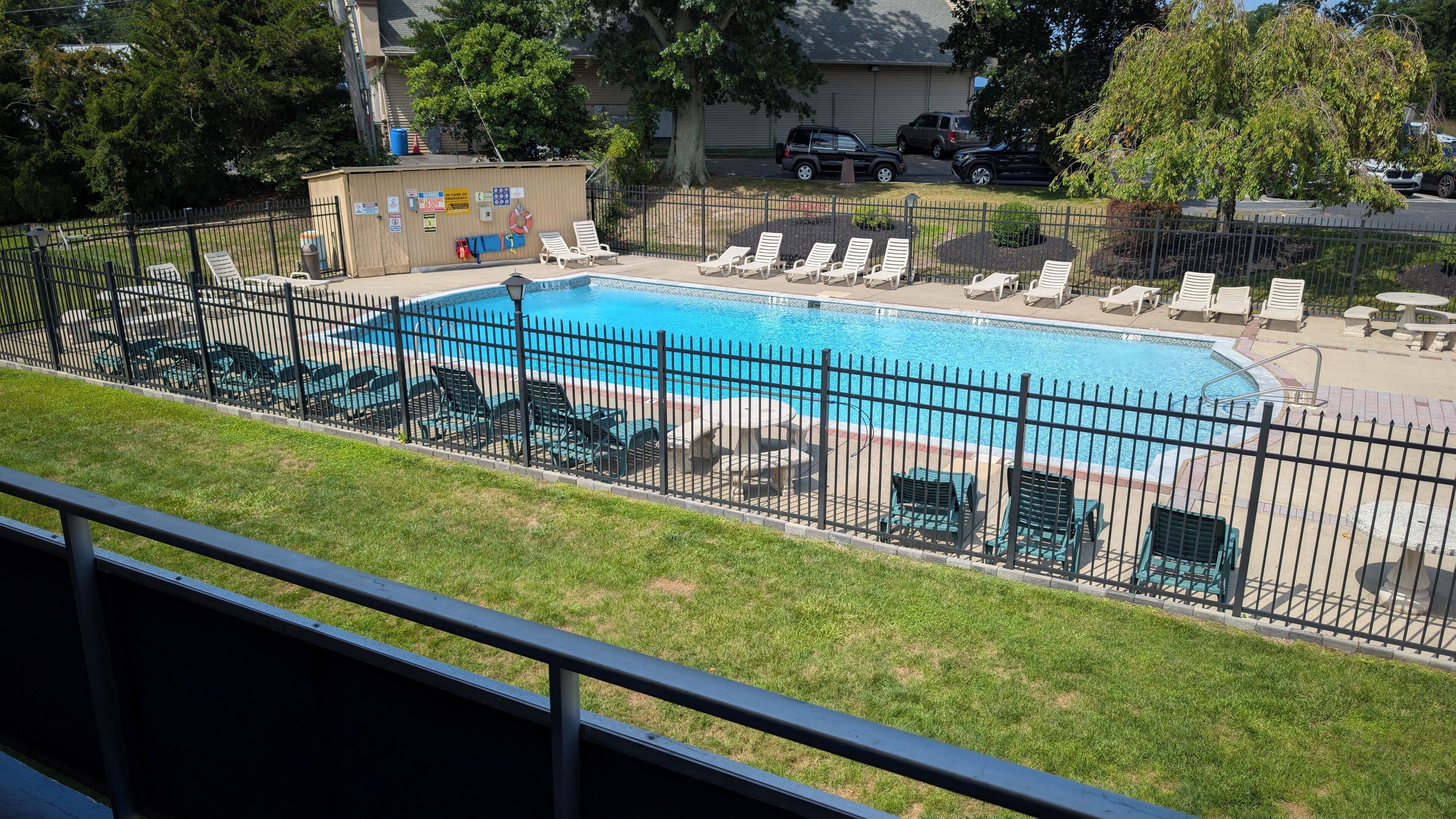 Piscina com espreguiçadeiras e área cercada, rodeada por grama e árvores.