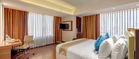 Quarto de hotel com cama, escrivaninha, cadeira e televisão.