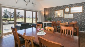 Uma sala de jantar com mesa e cadeiras de madeira, papel de parede floral e vista para um parque através de grandes janelas.
